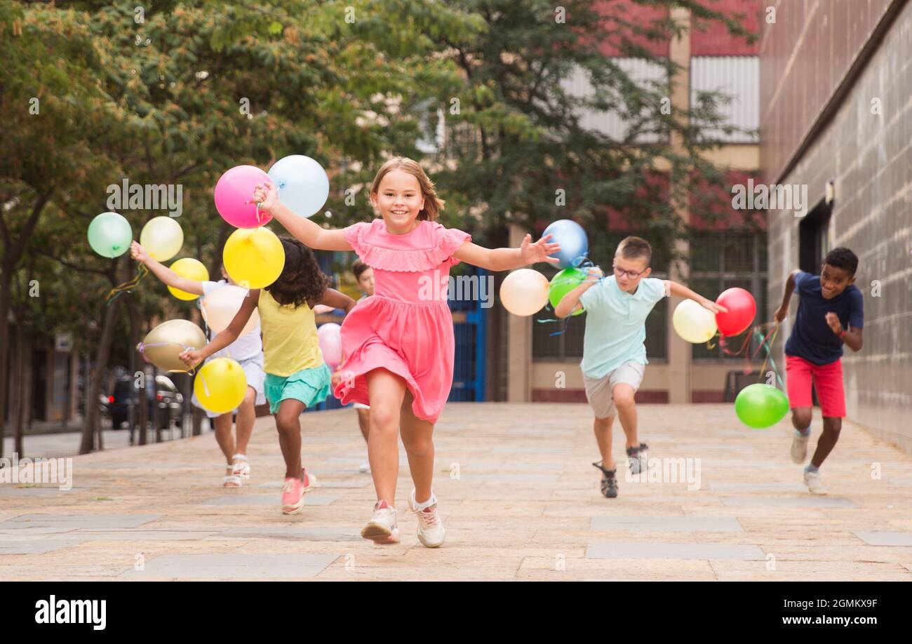 Happy tweens wth colorful balloons chasing each other on city street ...