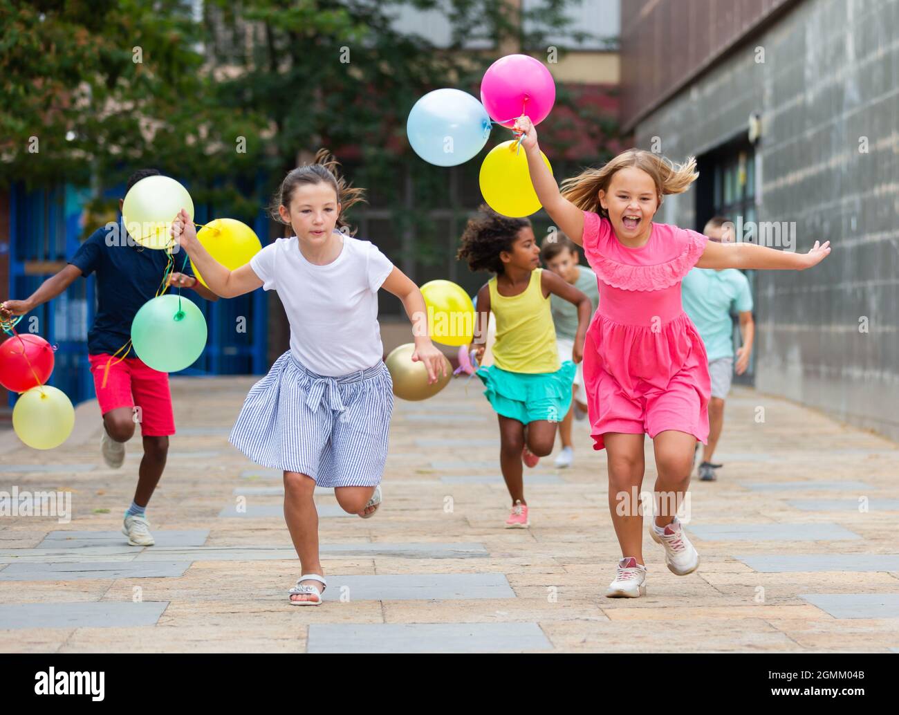 Happy tweens wth colorful balloons chasing each other on city street ...