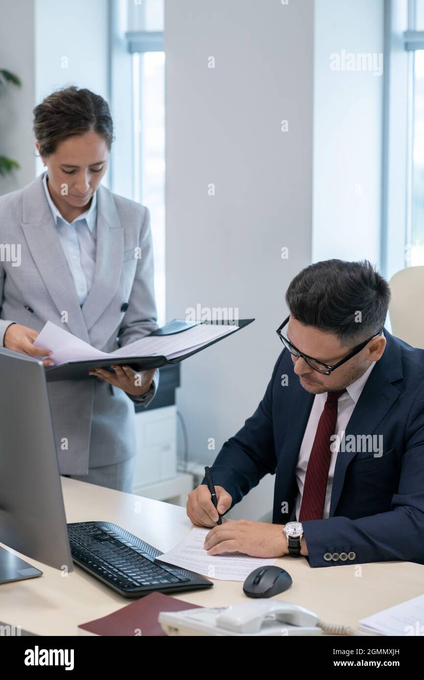 Mature employer signing documents by workplace while his secretary ...