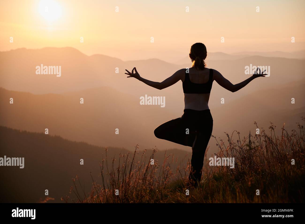 Back view of young woman performing yoga pose on grassy hill with ...