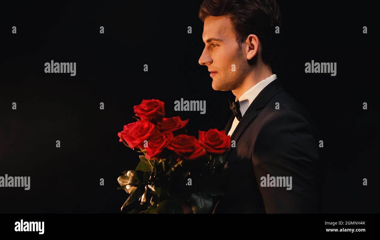 Side view of elegant young man holding red roses isolated on black ...