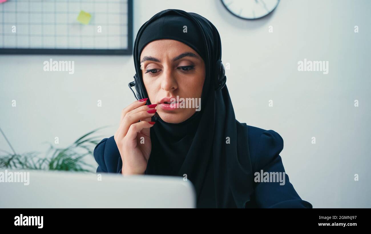 Muslim young woman in hijab using headset near blurred laptop in call ...