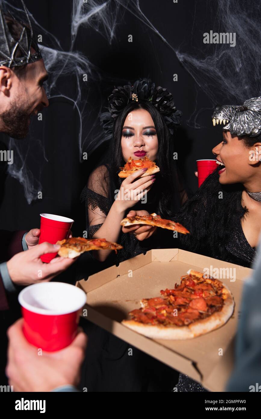 pleased asian woman in vampire halloween costume eating pizza near ...