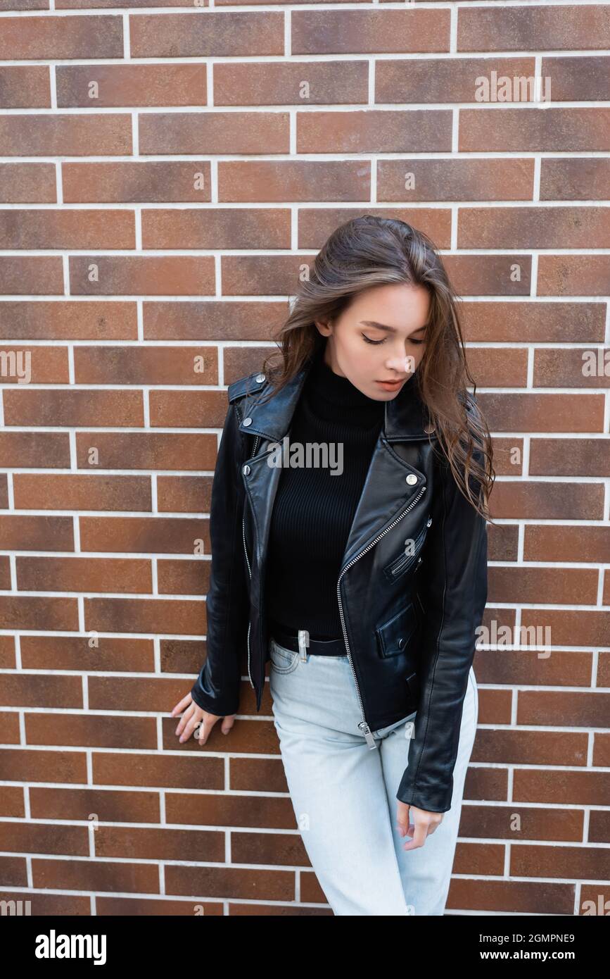 Pretty Young Woman In Leather Jacket And Jeans Standing Near Brick Wall