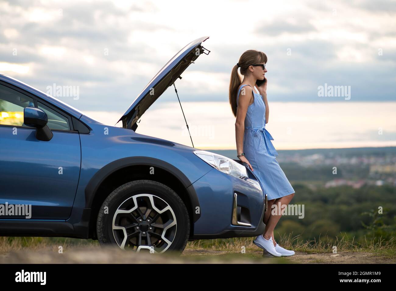 Young upset woman driver talking on mobile phone near a broken car with open hood inspecting ...