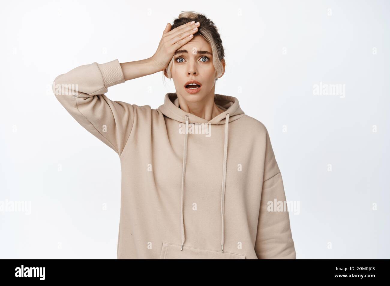 Worried young woman looking alarmed at camera, hold hand on head ...