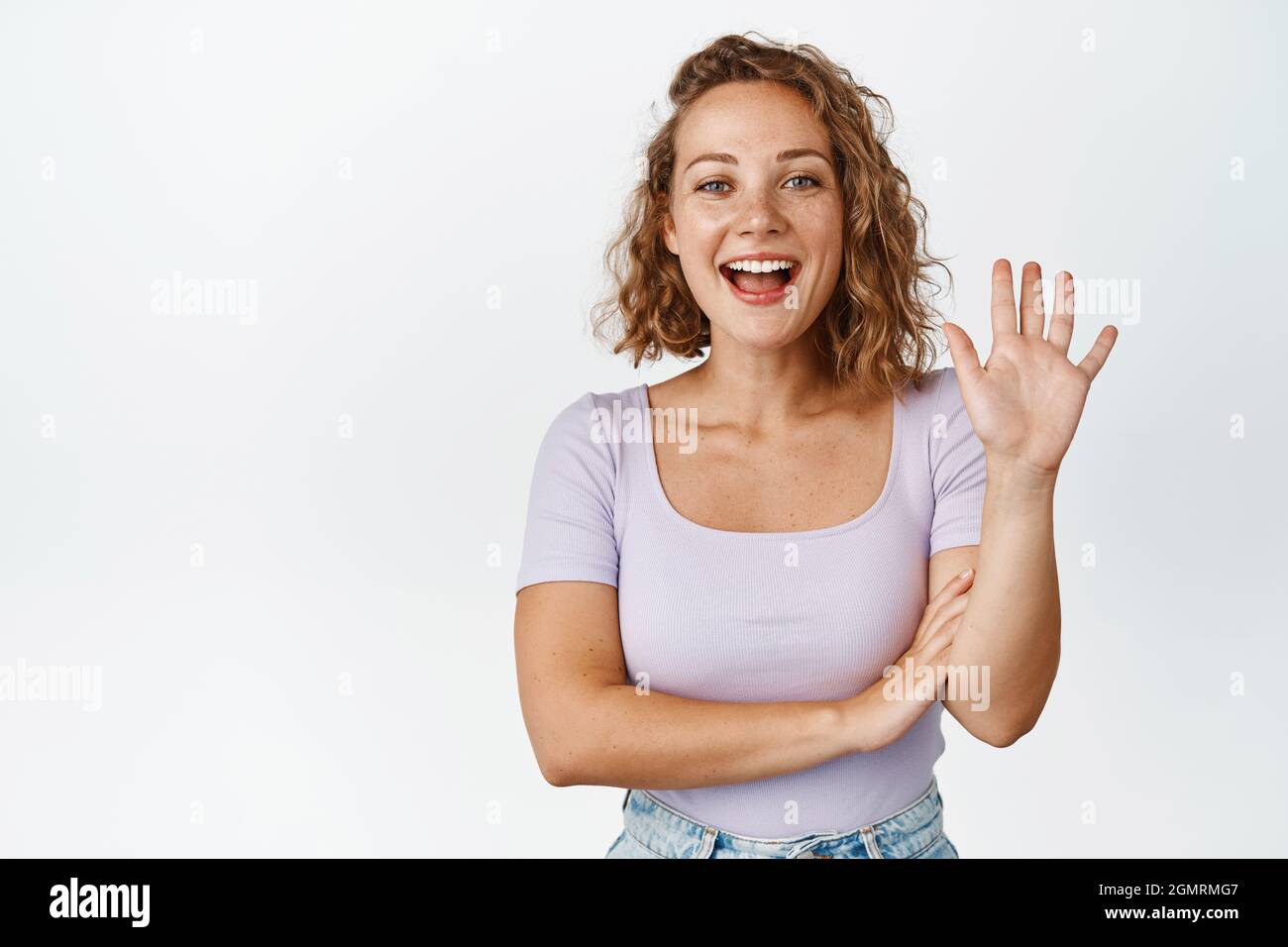 Friendly blond girl saying hi, waving raised hand and smiling at camera ...
