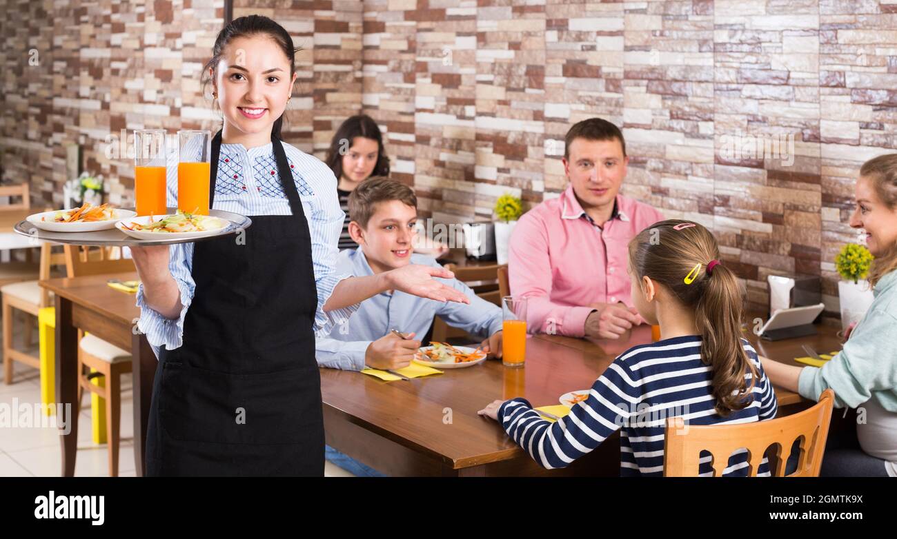 Attractive waitress holding tray with dishes meeting visitors in cafe ...