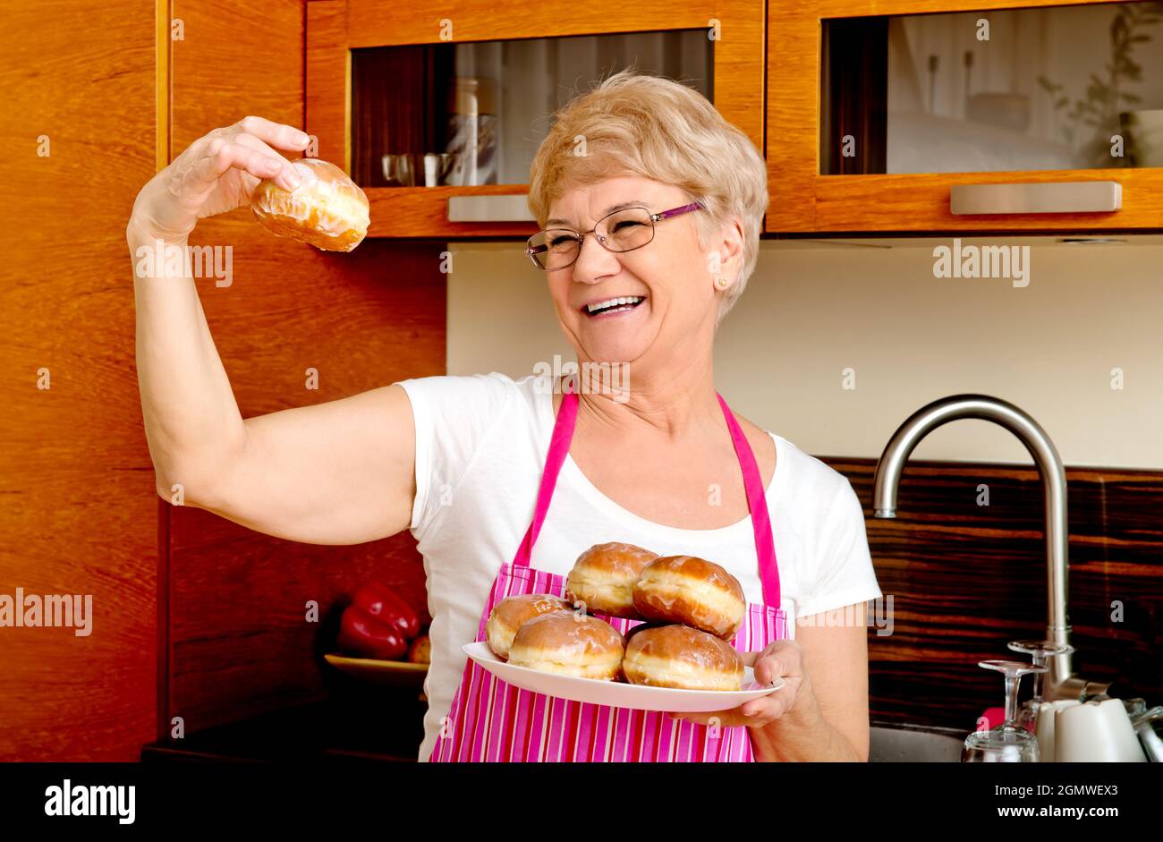 Cheerful elderly Polish woman showing homemade doughnuts in the kitchen ...