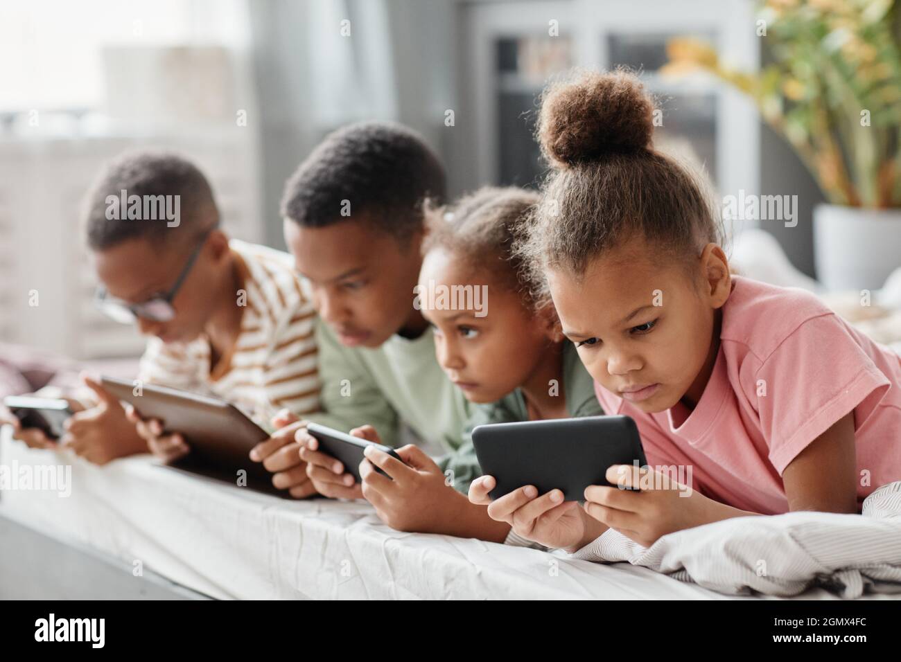 Four African-American kids using gadgets in row while lying on bed ...