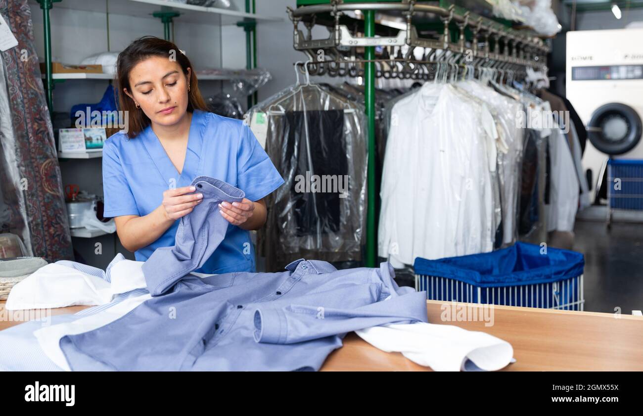 Focused female dry cleaning employee examining clean clothes before ...