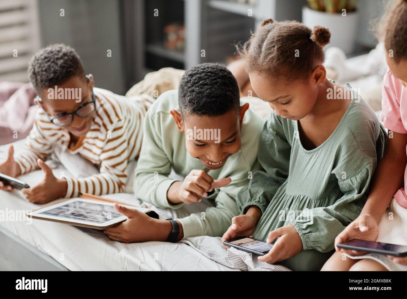 Happy African-American children using gadgets in row while lying on bed ...