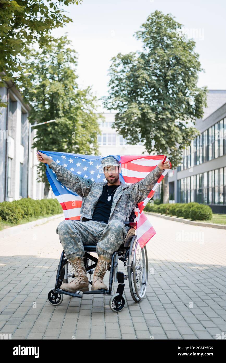 Young Disabled Veteran In Wheelchair Holding Usa Flag On Urban Street young-disabled-veteran-in-wheelchair-holding-usa-flag-on-urban-street