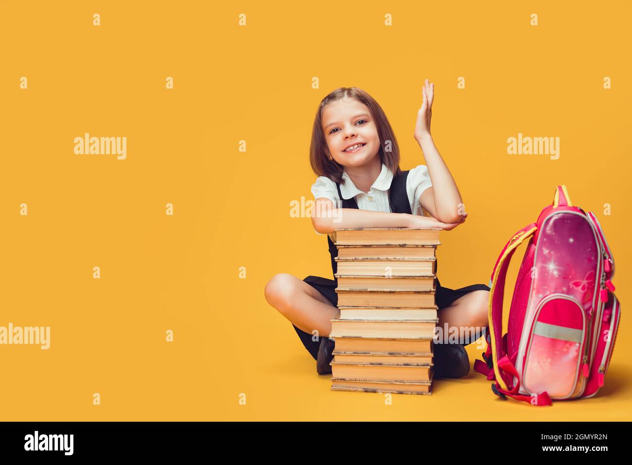 Smiling schoolgirl sitting behind a stack of books and raising hand ...
