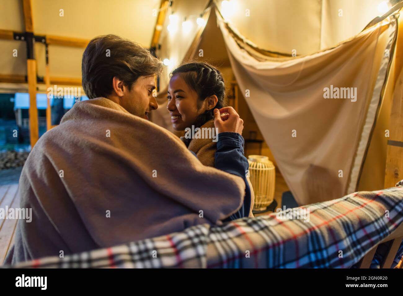 Man in blanket touching smiling asian girlfriend on bench near glamping ...