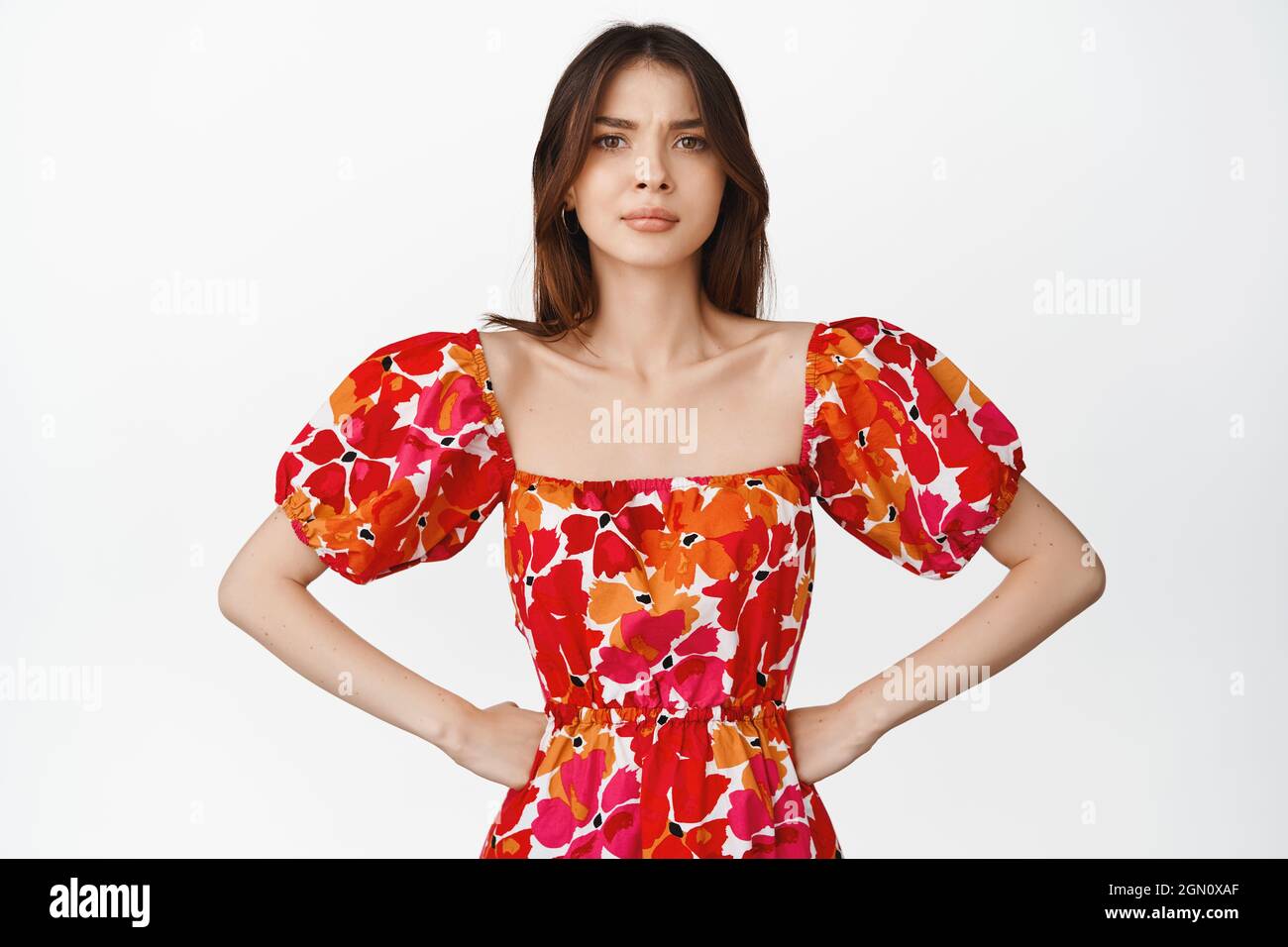 Young bothered girl in red dress, holding hands on waist, frowning and ...