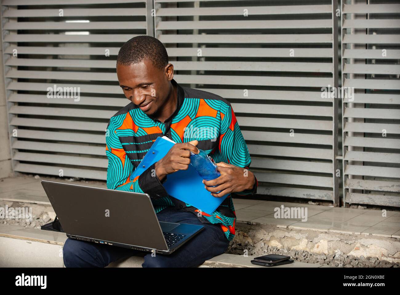 happy young african man sitting in the street and working on the laptop ...