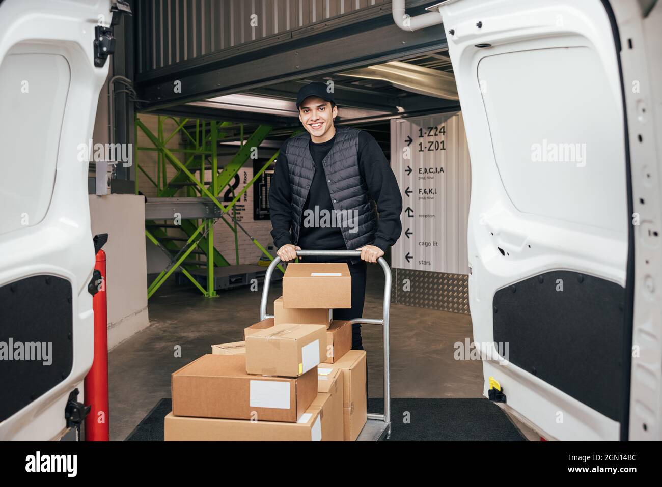 Smiling courier in uniform standing in warehouse with cart preparing ...