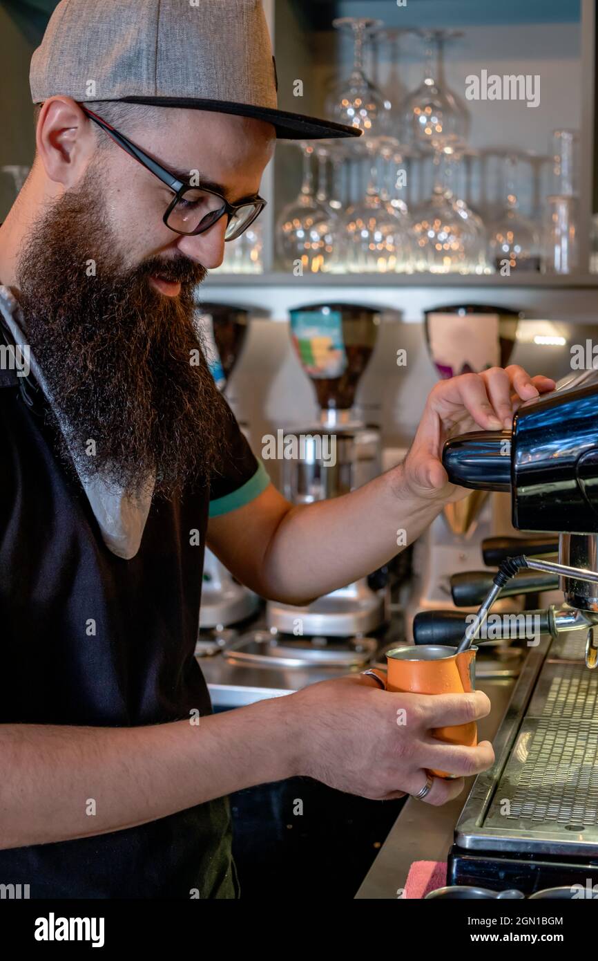 Bearded barista making steamed milk cream in a steel jug under a steam ...