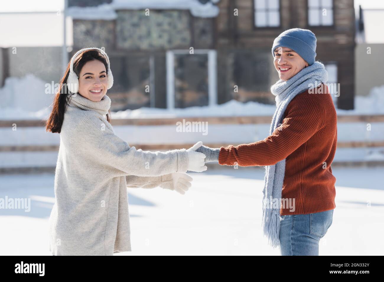 smiling young woman in ear muffs holding hands with cheerful boyfriend ...