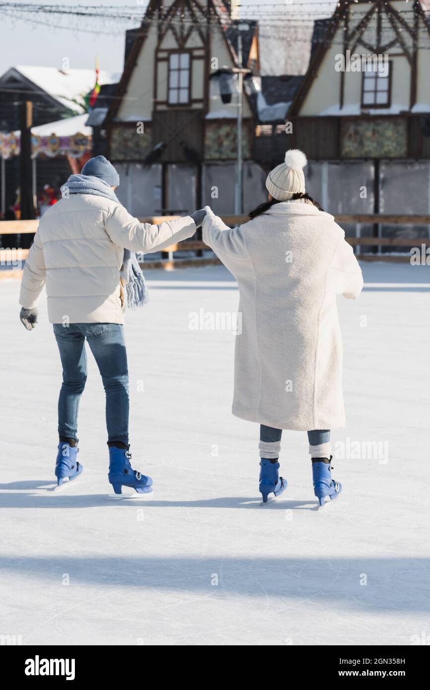 back view of young man and woman in ice skates holding hands on ice ...