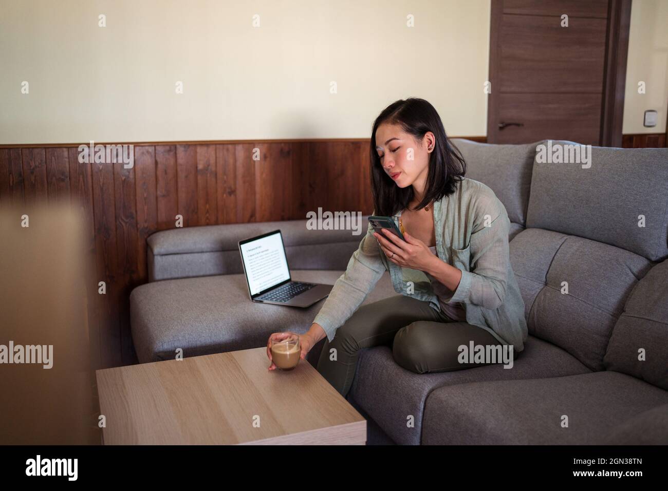 Young ethnic female with glass of coffee surfing internet on cellphone ...