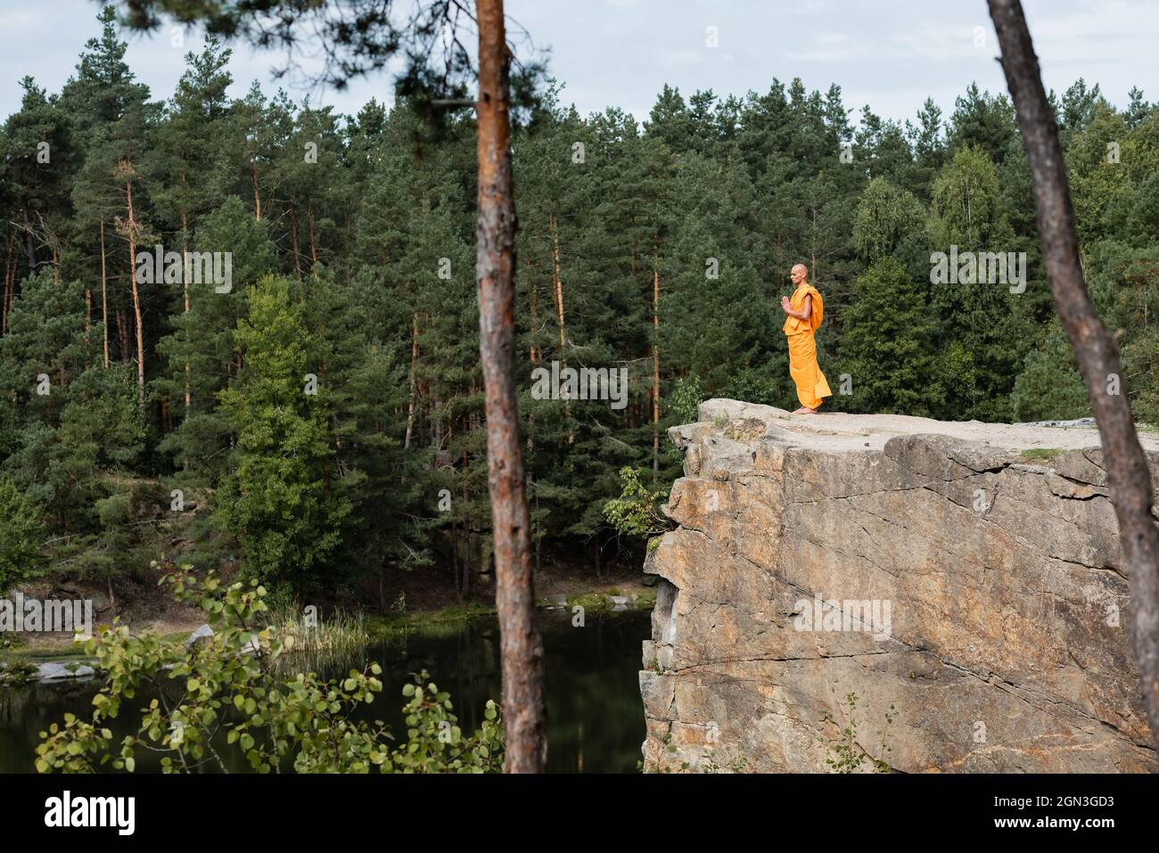 buddhist monk in orange kasaya praying on high rocky cliff over river ...