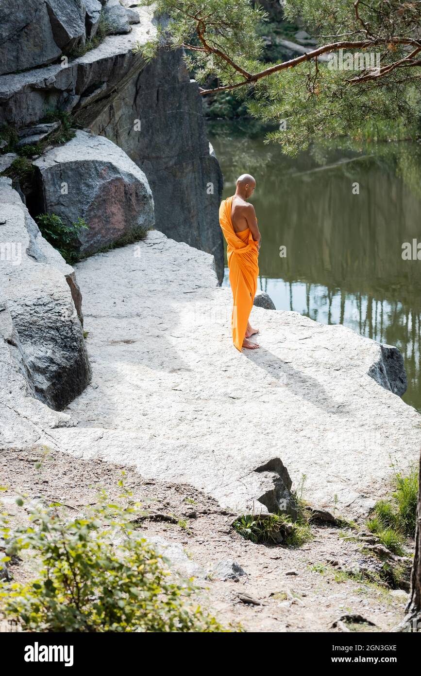 high angle view of buddhist monk meditating near rocks over forest lake ...