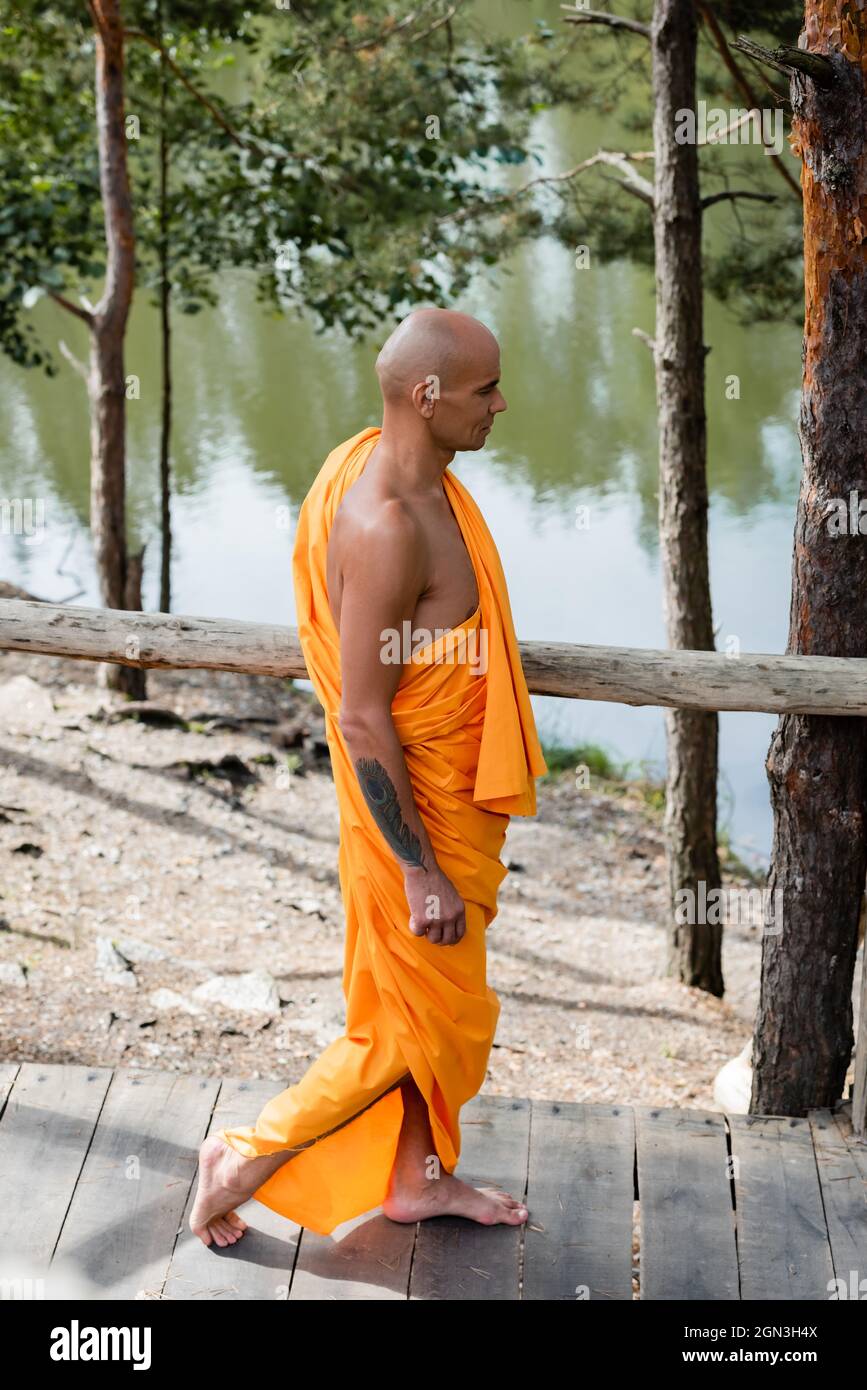 side view of buddhist monk walking on wooden walkway near forest lake ...