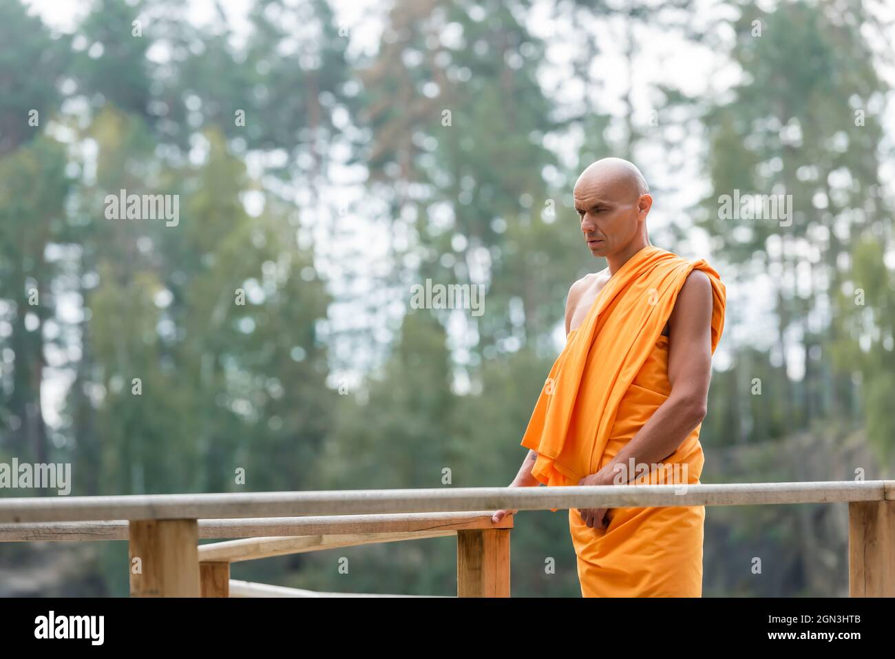 man in traditional buddhist robe standing near wooden fence in forest ...