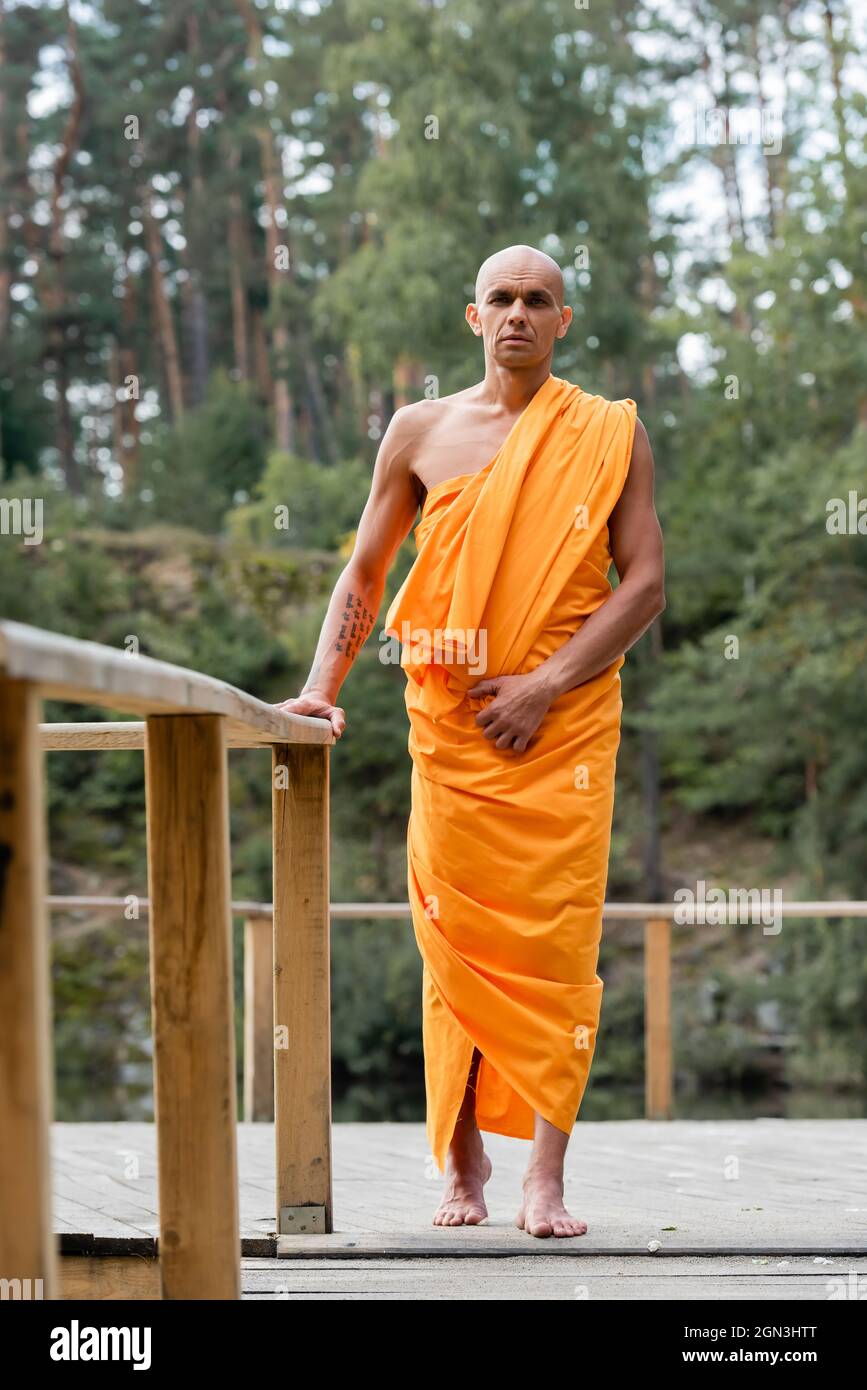 full length view of buddhist monk standing near wooden fence in forest ...