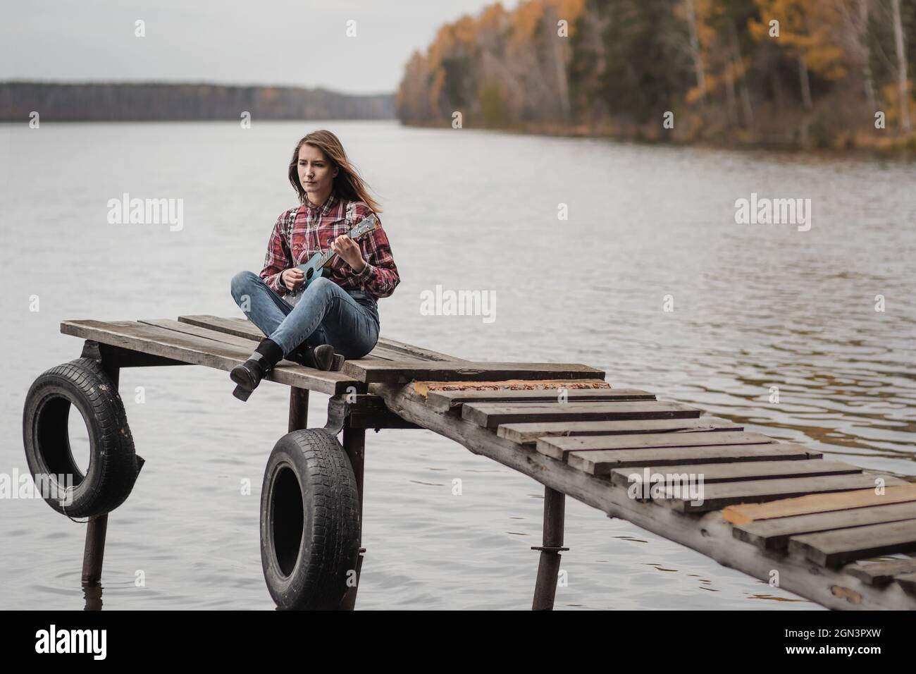 Young woman sitting and playing ukulele guitar on wooden pier in autumn ...