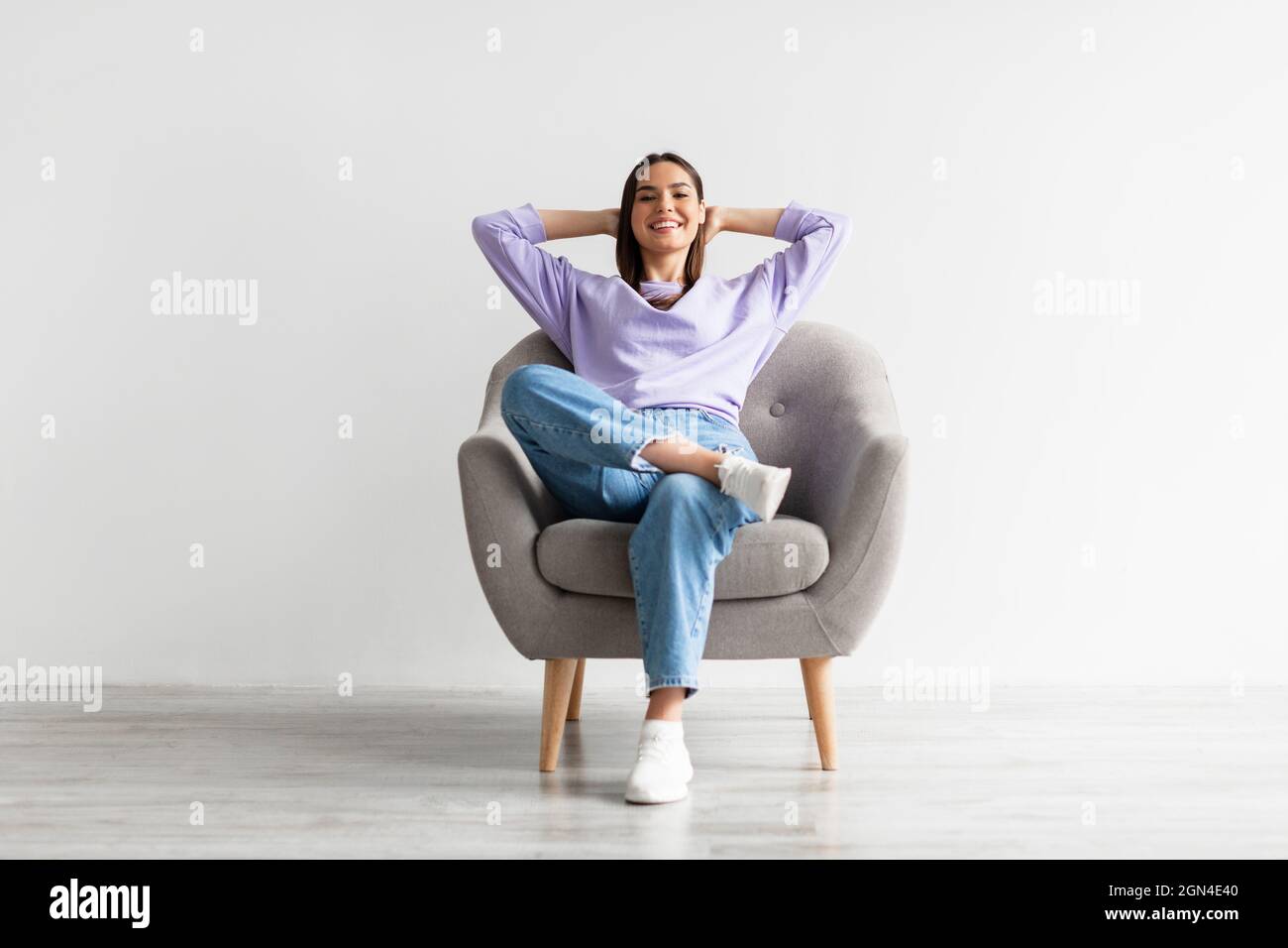 Happy young woman sitting in armchair with hands behind head, resting ...
