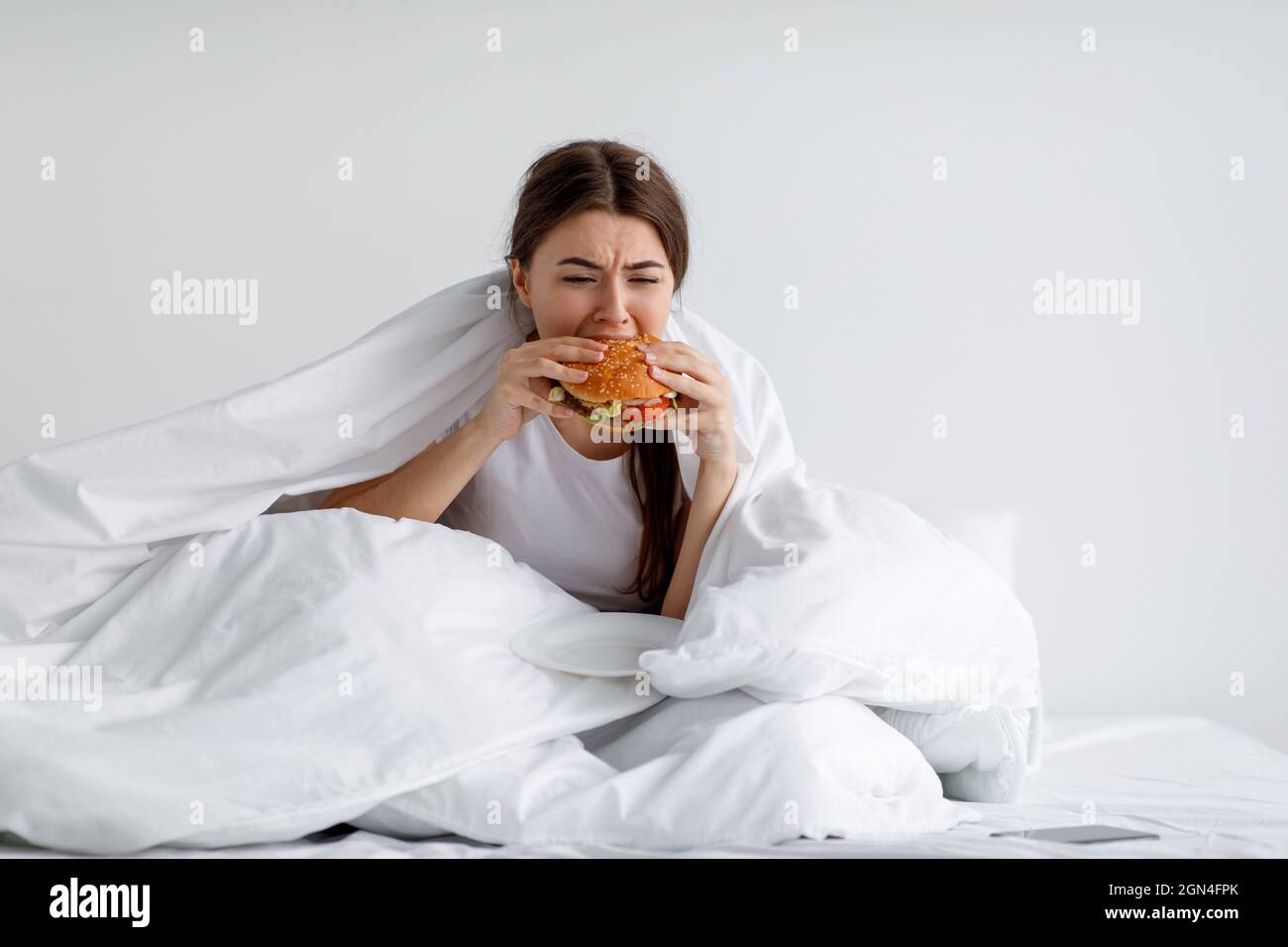 Hungry young female greedily eating burger on bed. Eating disorder ...