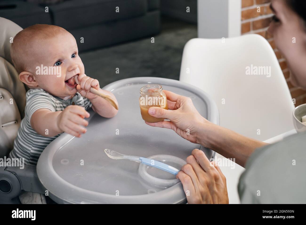 Mother giving food to baby boy, pure from spoon in highchair at home ...