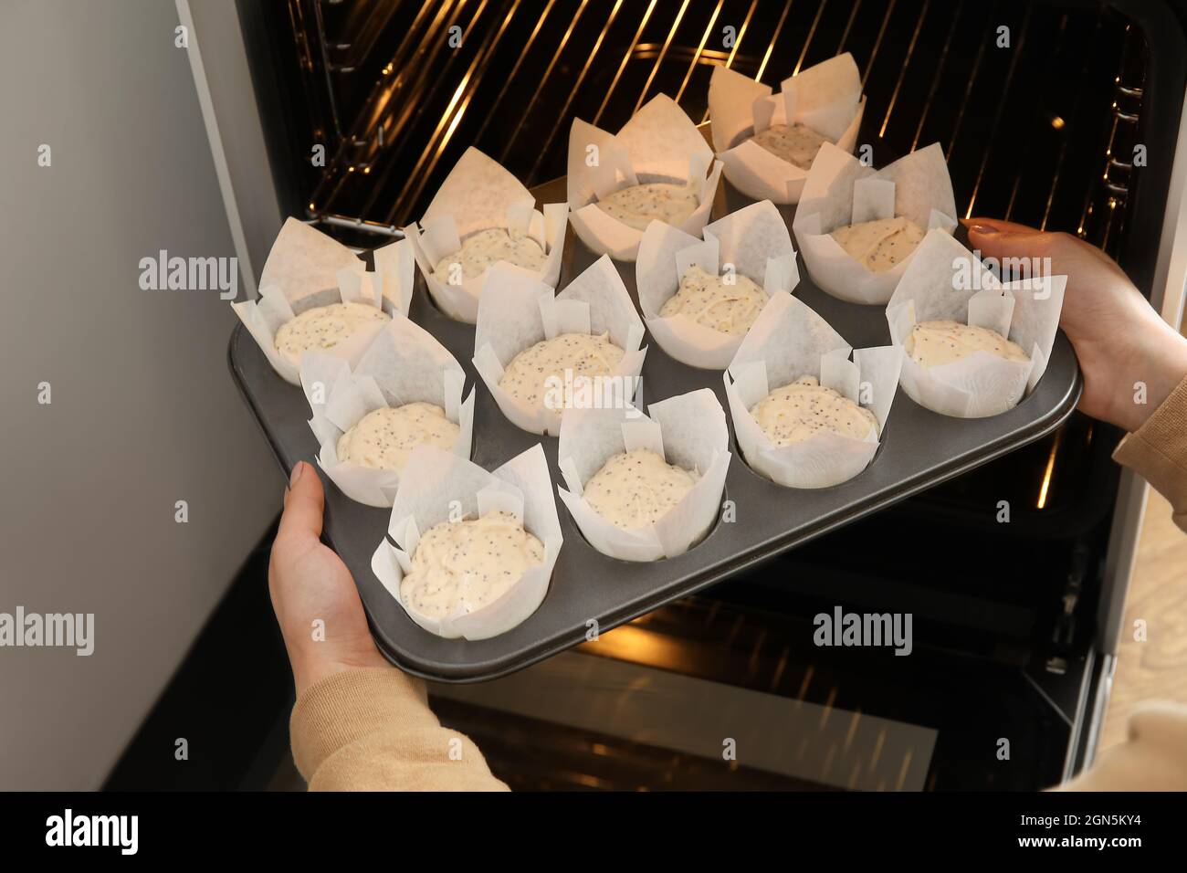 Woman putting uncooked muffins with poppy seeds to oven in kitchen ...