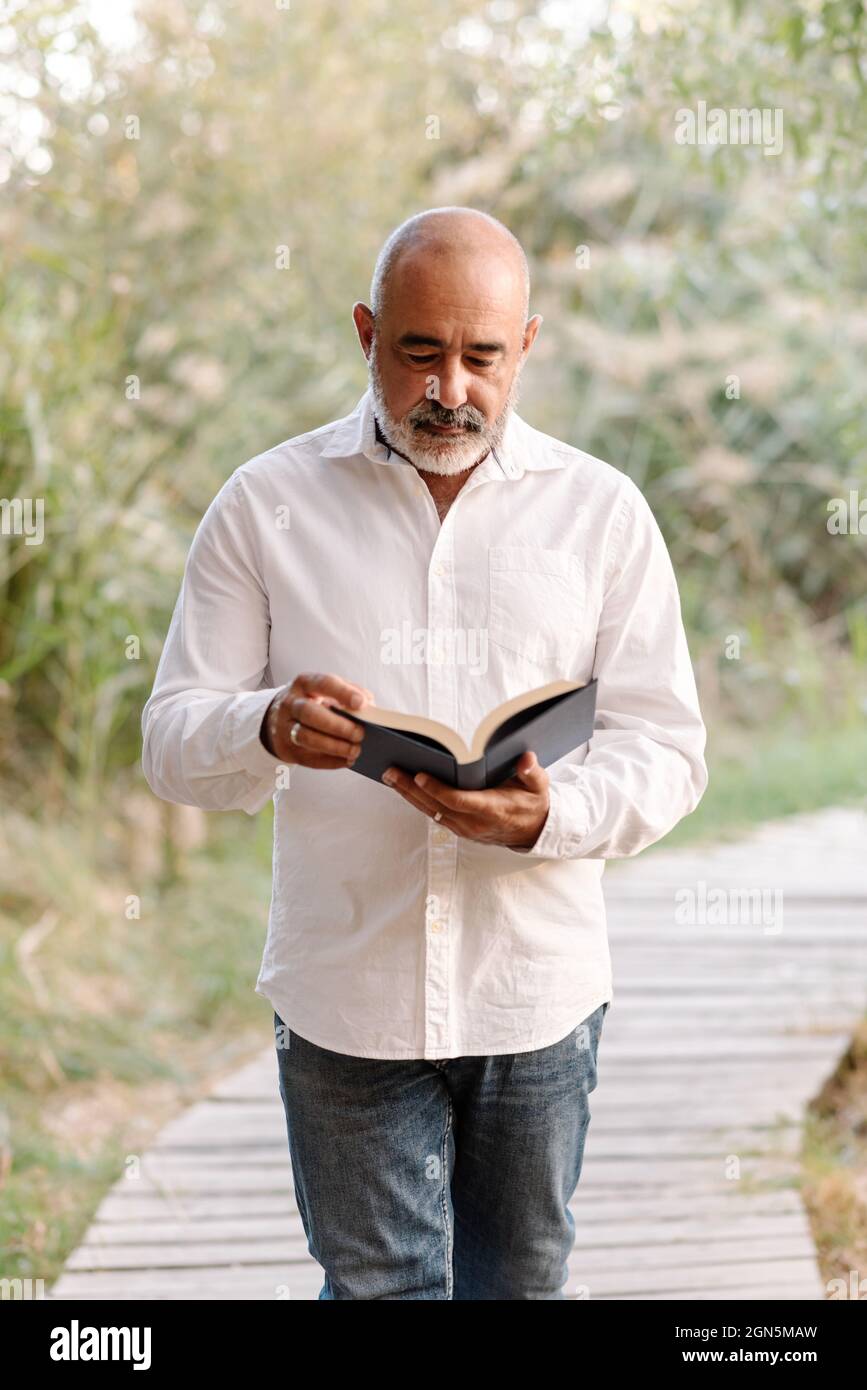 Concentrated adult man reading a book while standing outdoors in a park ...