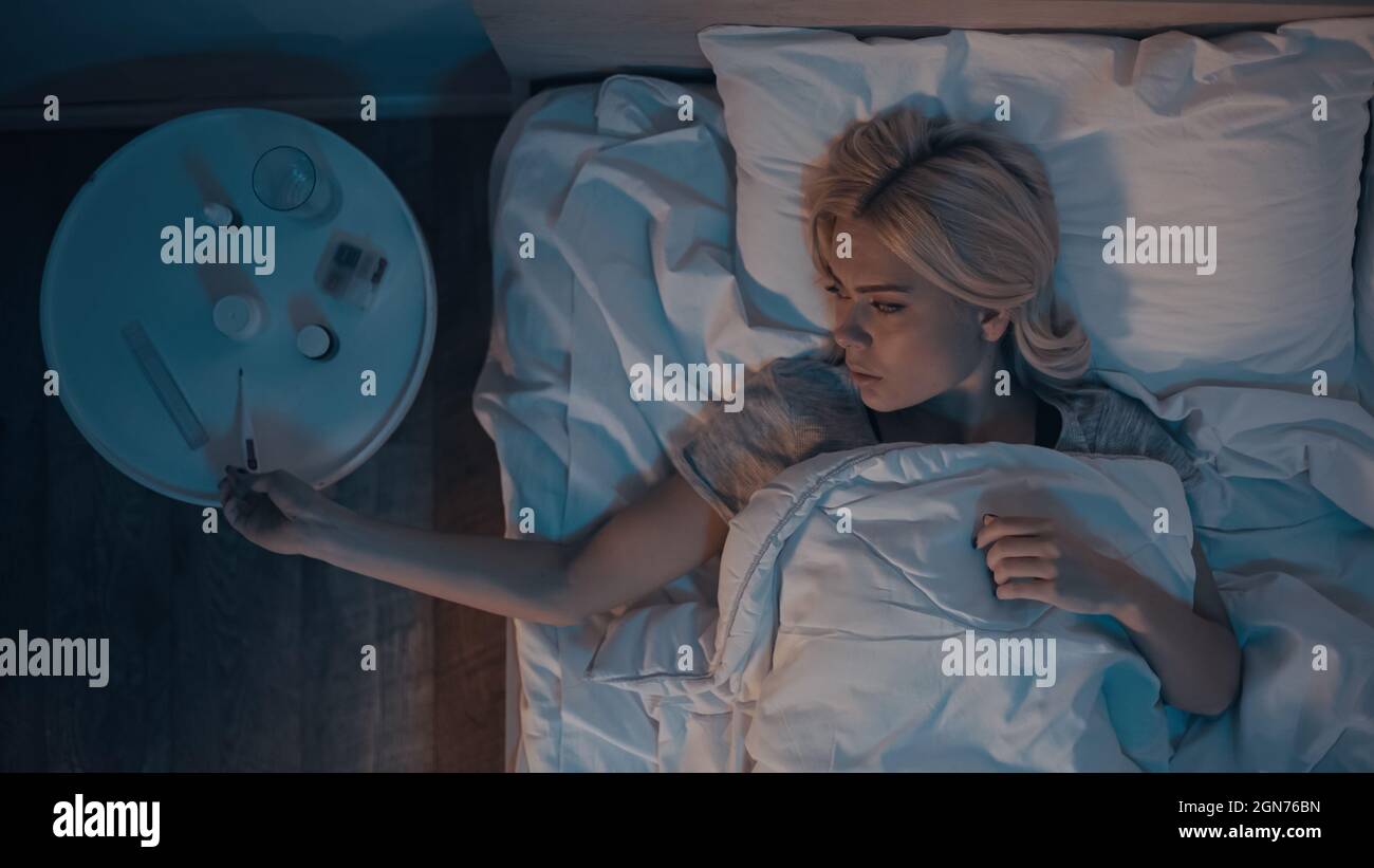 Top view of sick woman looking at thermometer on bedside table in ...