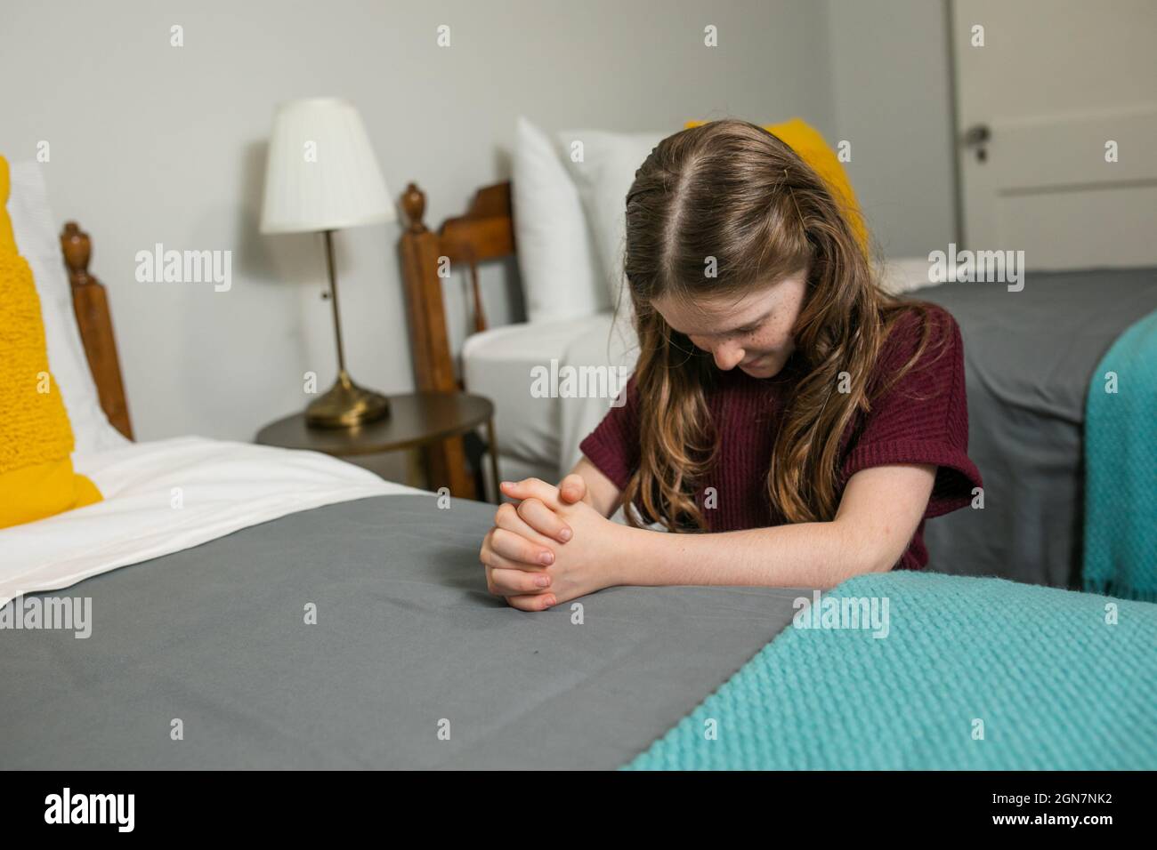 A young girl kneeling and praying to God at the side of a bed in her ...