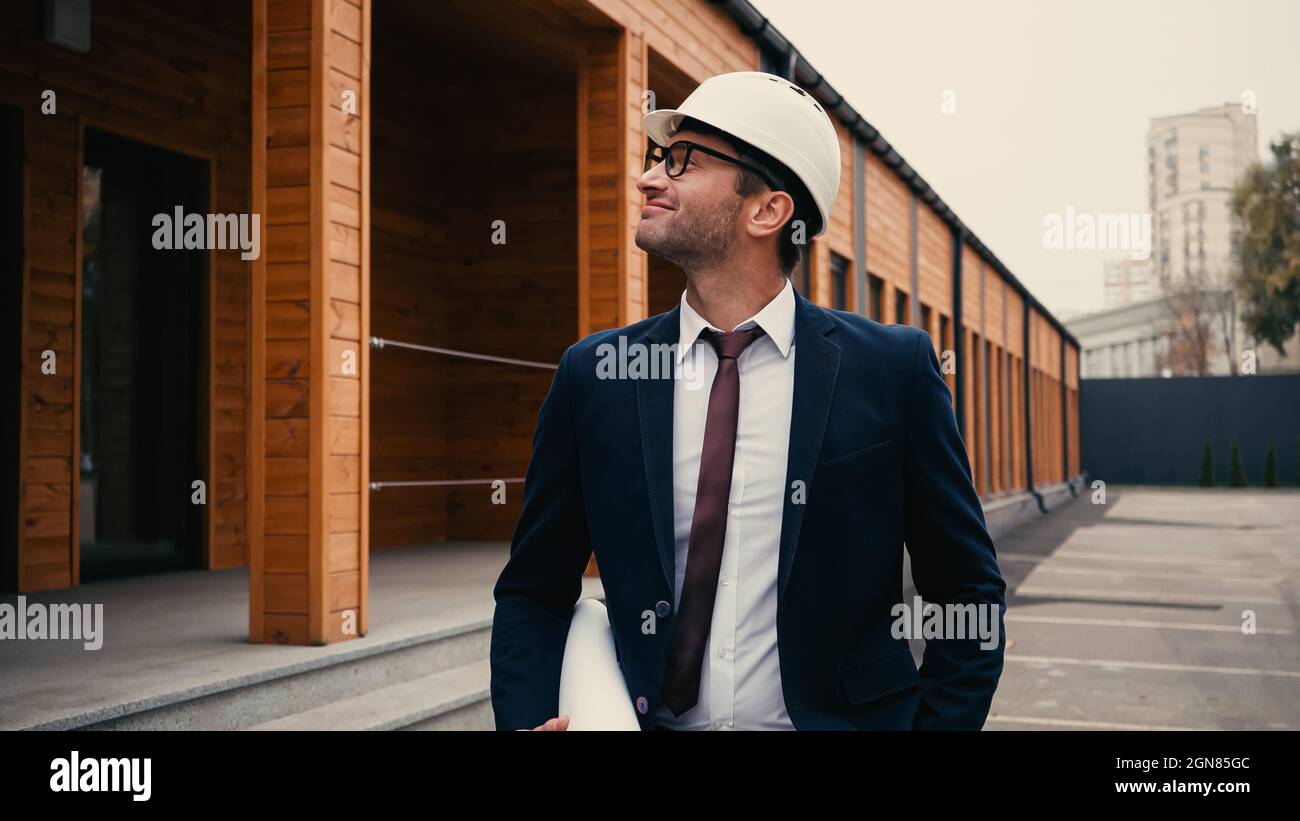 Side view of smiling engineer in hardhat holding blueprint and looking ...