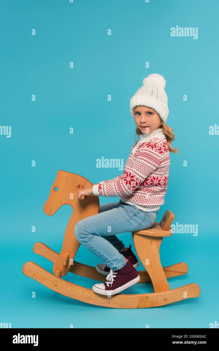 girl in warm hat and sweater riding rocking horse while looking at ...