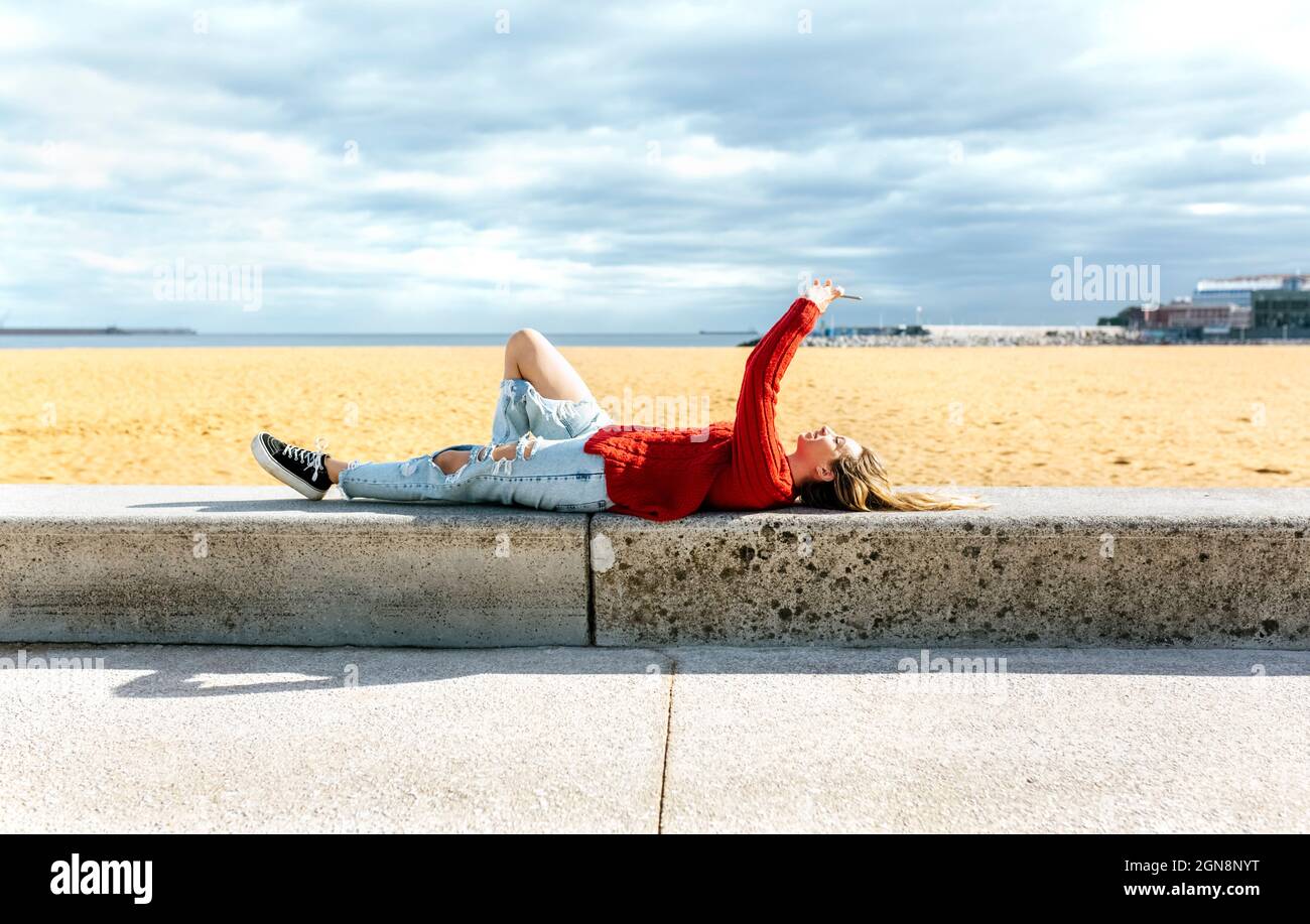 Happy woman using smart phone while lying down on retaining wall at ...