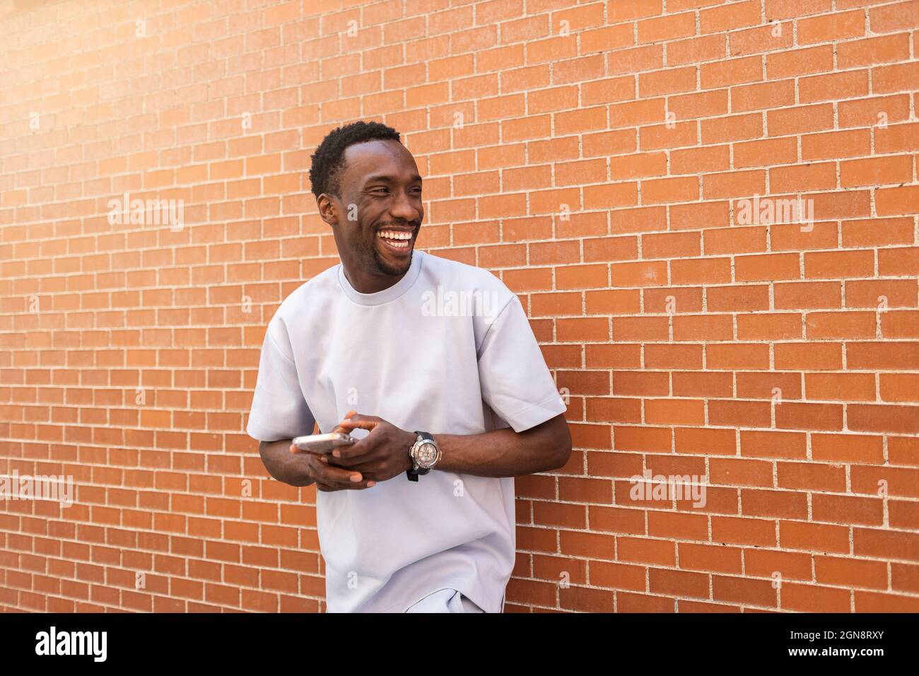 Young man laughing while holding smart phone in front of brick wall ...