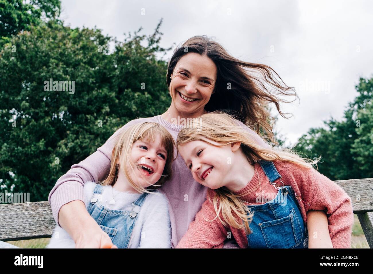 Cheerful mother with arms around daughters sitting on bench Stock Photo ...