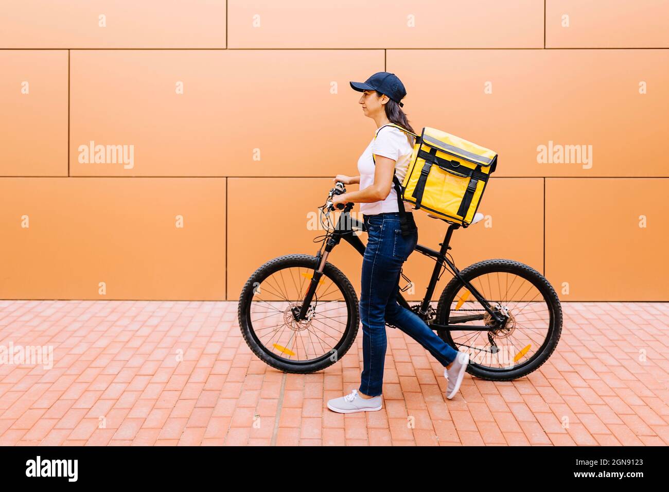 Delivery woman with backpack wheeling bicycle on footpath Stock Photo ...