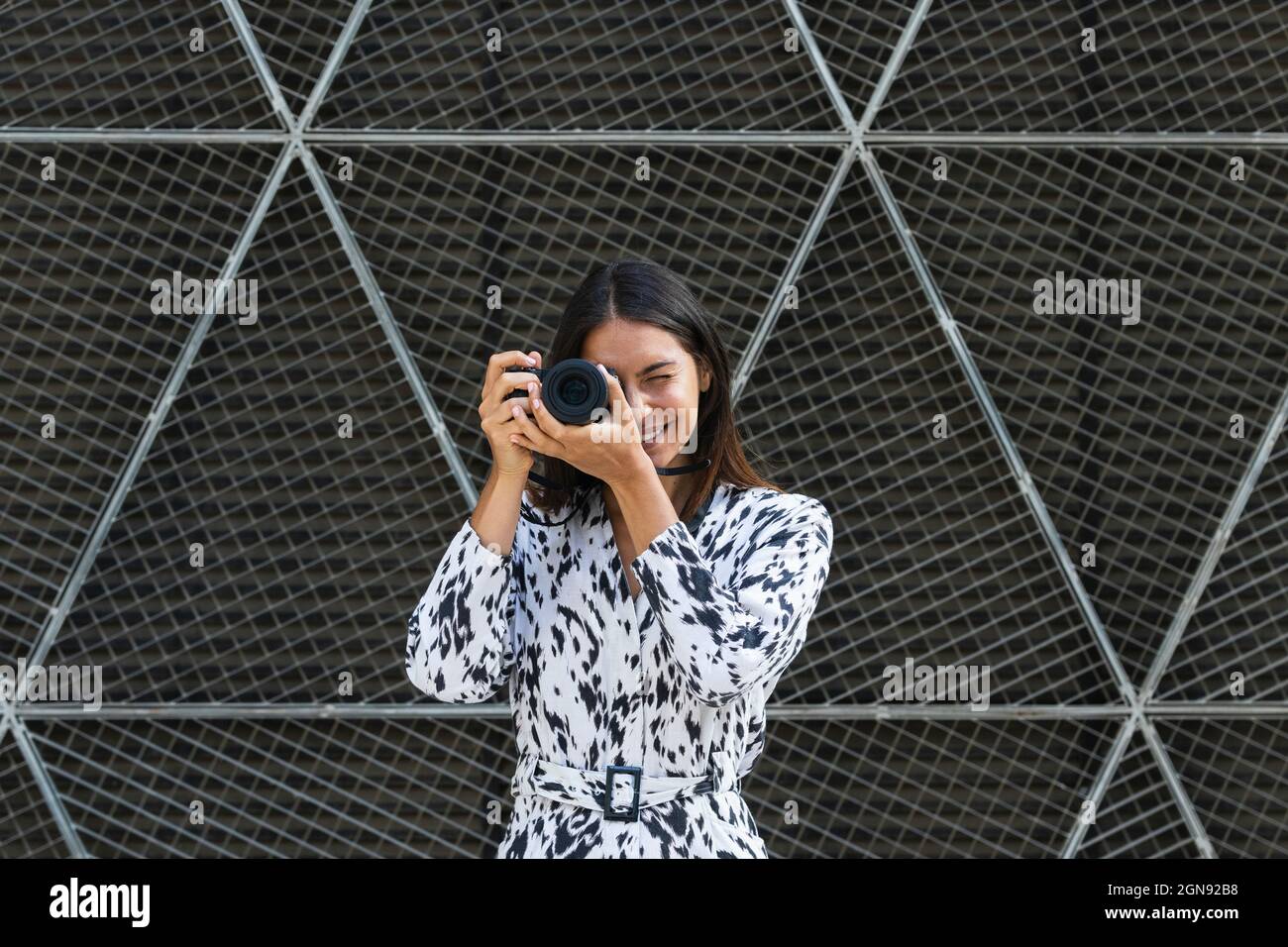 Smiling young woman photographing through camera in front of fence ...