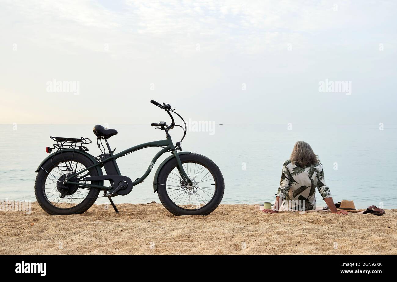 Mature man sitting by bicycle while relaxing at beach Stock Photo - Alamy