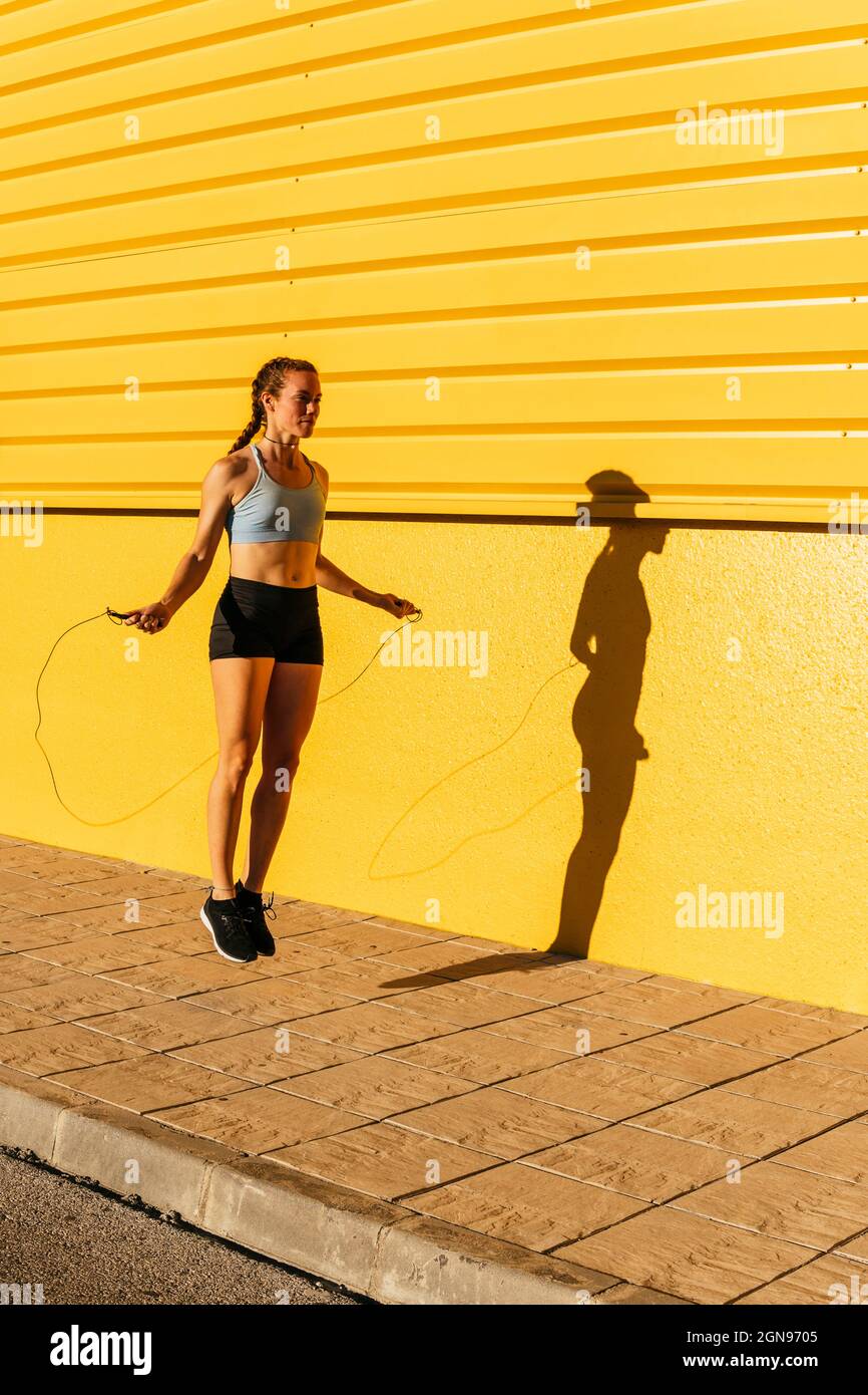 Female athlete using jump rope while practicing skipping by yellow wall ...