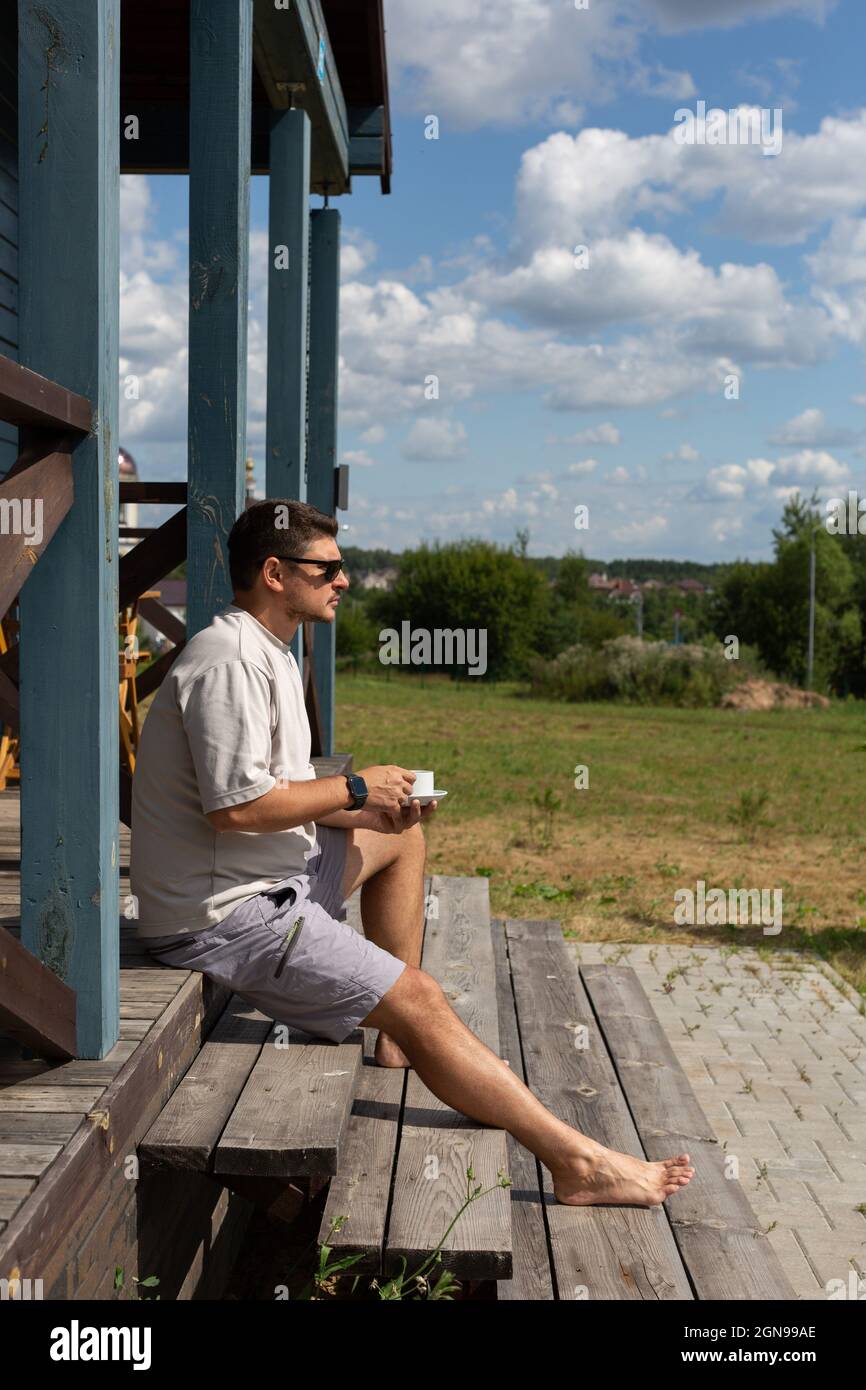A young man is sitting on the porch of a house with a cup of coffee ...