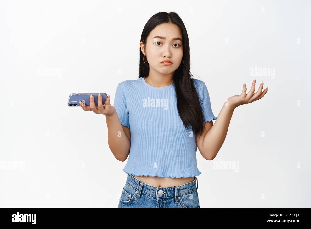 Confused asian girl holding smartphone and shrugging shoulders ...